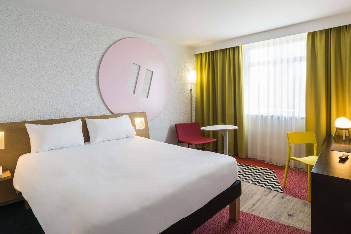 Quarto de hotel com uma cama grande, uma escrivaninha, uma cadeira e uma mesinha.