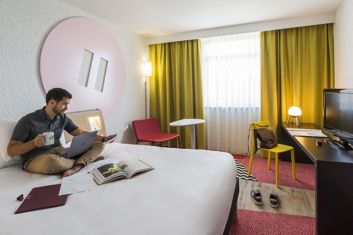 Quarto de hotel com cama, escrivaninha, cadeira, televisão e abajur.
