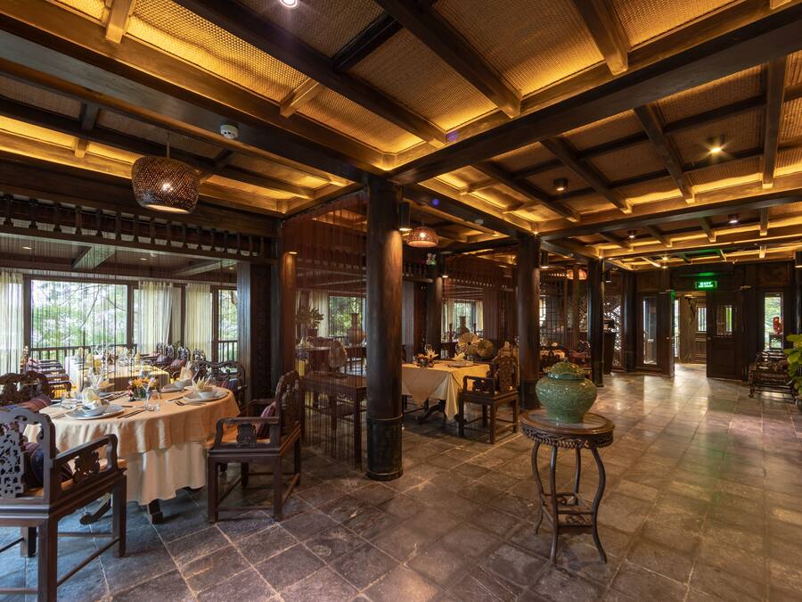 Uma ampla sala de jantar com pé direito alto e vigas de madeira, com mesas e cadeiras redondas e grandes janelas que permitem entrada de luz natural.
