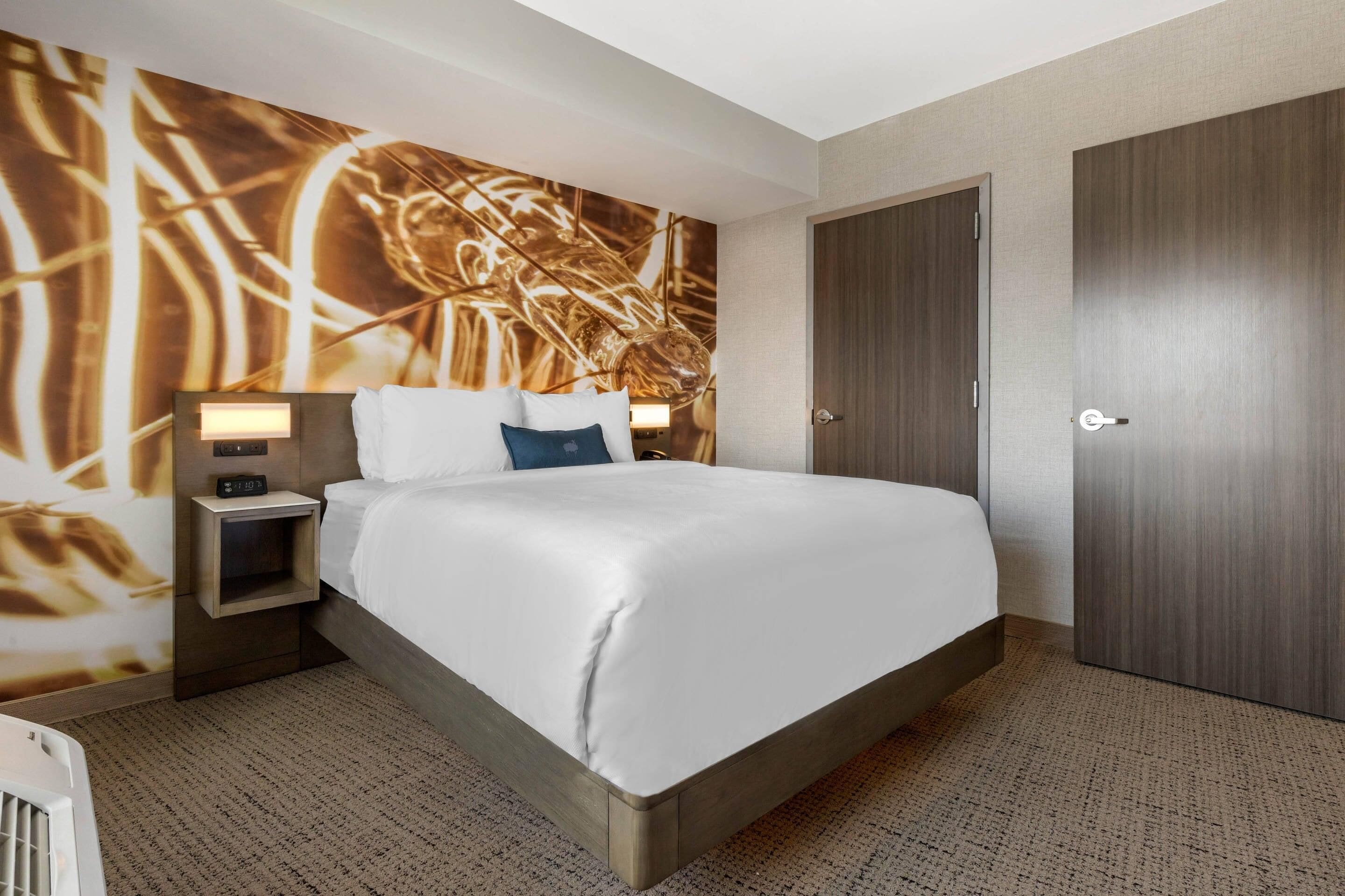 Quarto de hotel com uma cama grande, uma mesa de cabeceira, uma luminária na parede e uma porta.