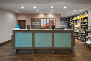 Balcão de recepção de hotel com um letreiro 'Welcome to Hampton', um quiosque de check-in digital e um bar de lanches com diversos alimentos.