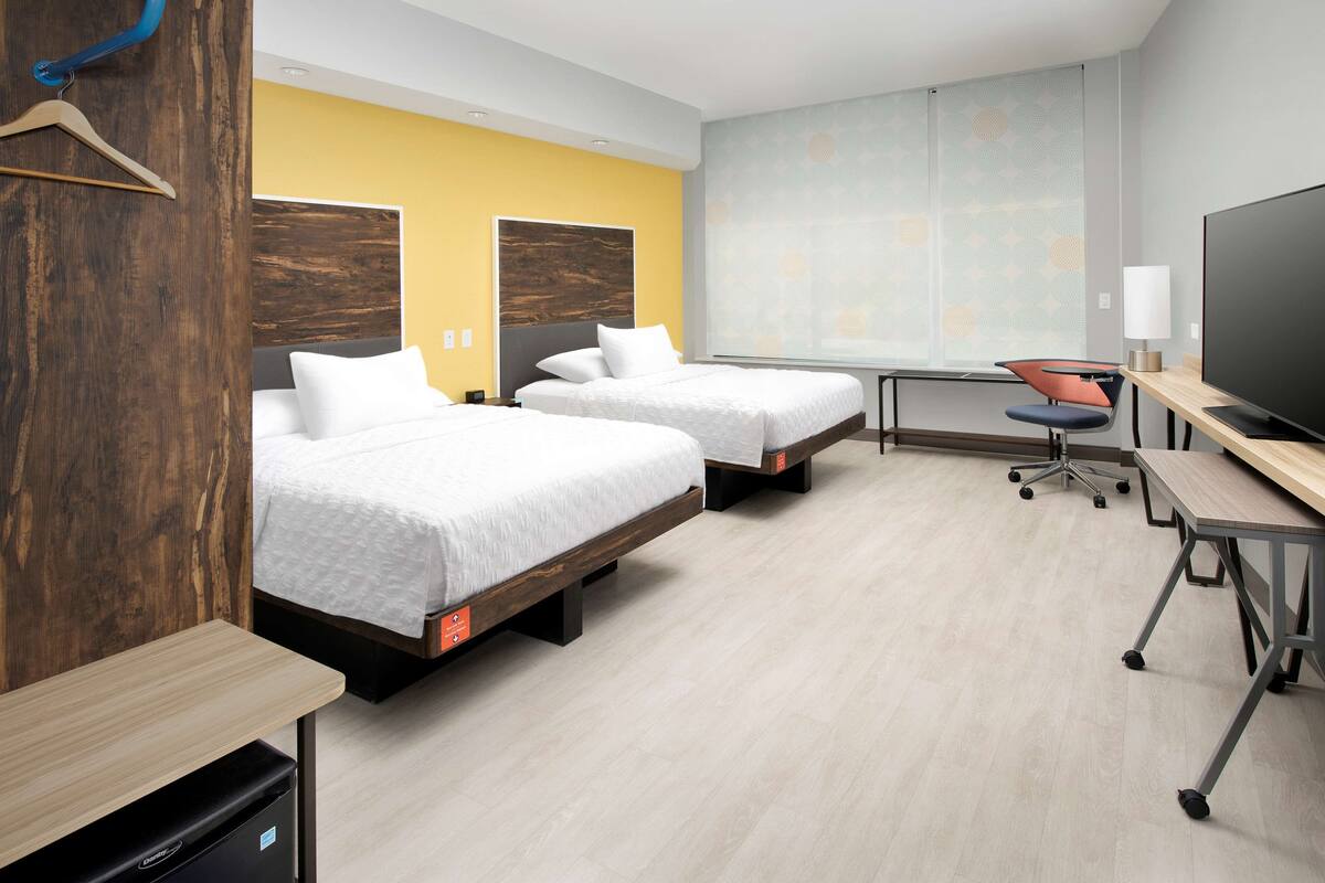 Quarto de hotel com duas camas, uma escrivaninha, uma cadeira, uma televisão e um guarda-roupa.