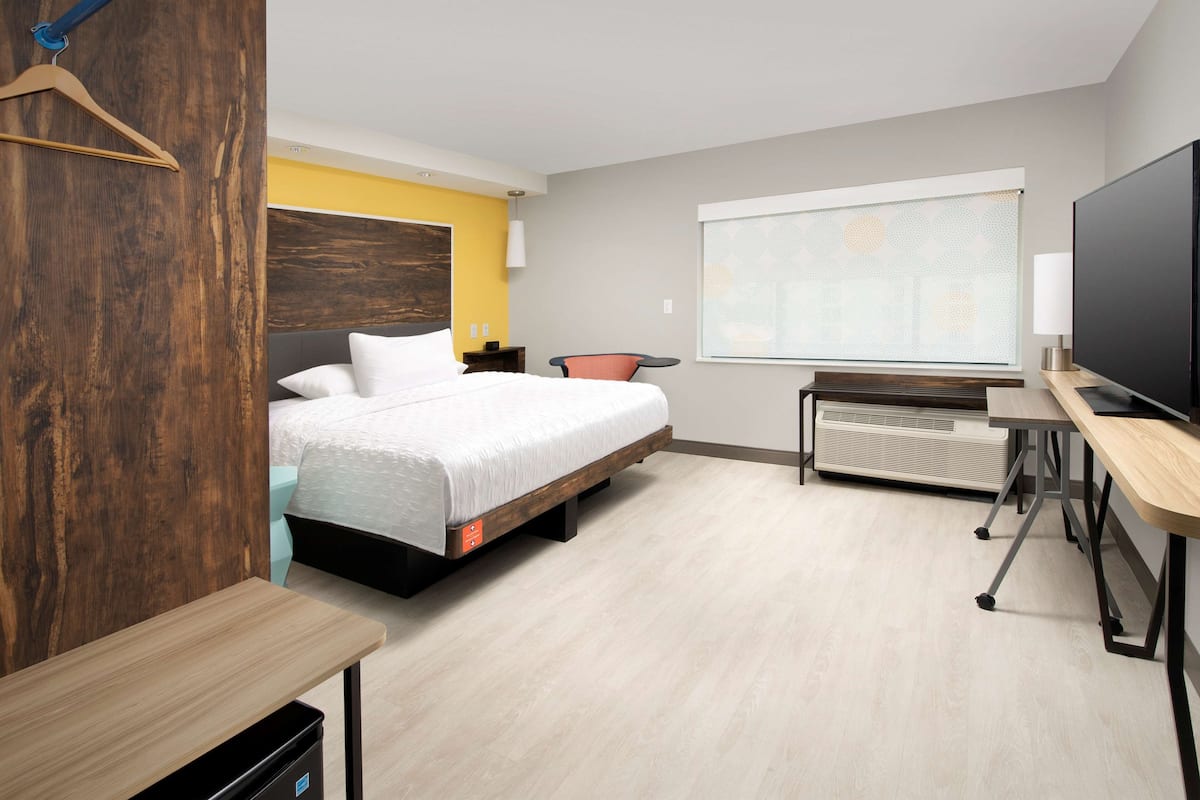 Quarto de hotel moderno com cama, escrivaninha, televisão e uma grande janela com persianas.
