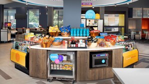 Uma estação de bebidas com serviço próprio, com variedade de lanches e bebidas, um refrigerador e um terminal de pagamento digital.