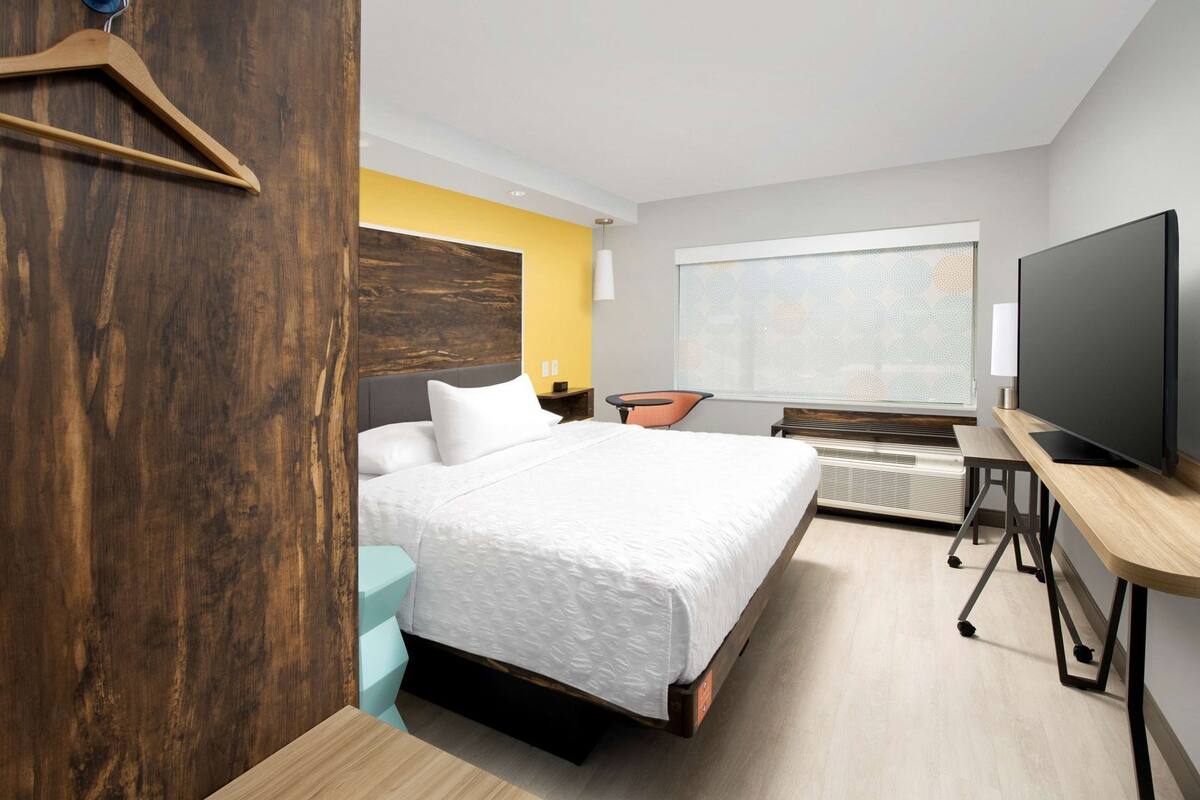 Quarto de hotel moderno com cama, escrivaninha, televisão de tela plana, cadeira e janela grande.