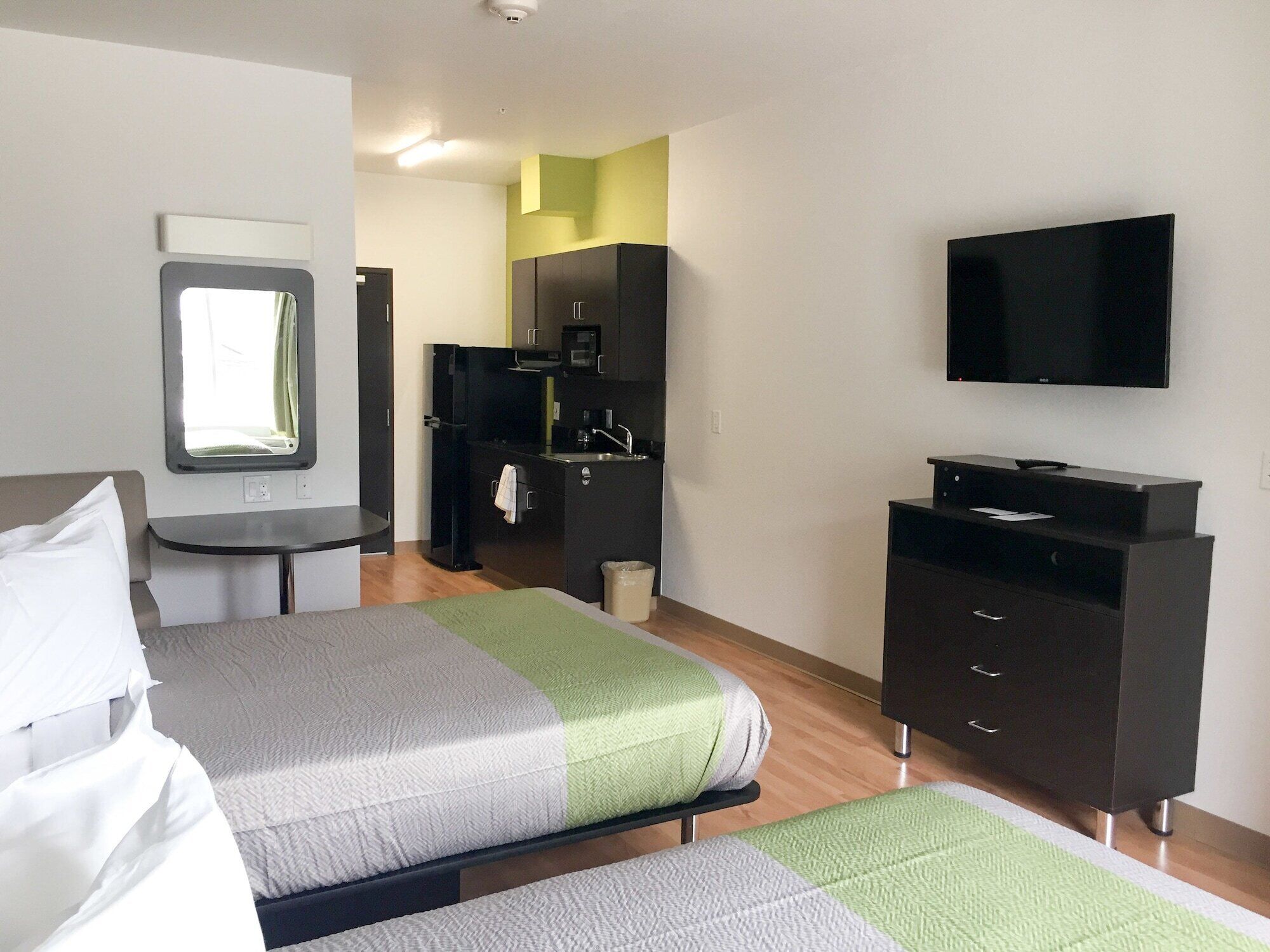 Quarto de hotel com cama, escrivaninha, televisão e cozinha compacta.