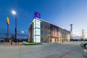 Um hotel com vários andares, um letreiro grande com o número '6' no telhado, janelas amplas e um estacionamento com vários veículos.