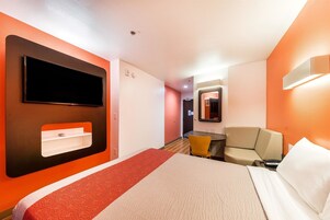 Quarto de hotel com uma cama, televisão na parede, mesa pequena, cadeira e espelho.