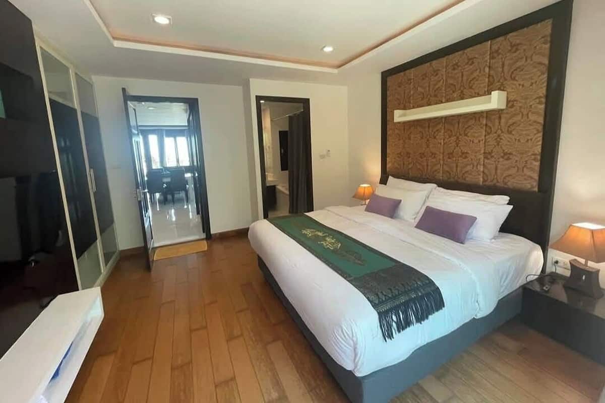 Quarto de hotel com uma cama grande, piso de madeira, espelho, escrivaninha e mesa de cabeceira com abajur.