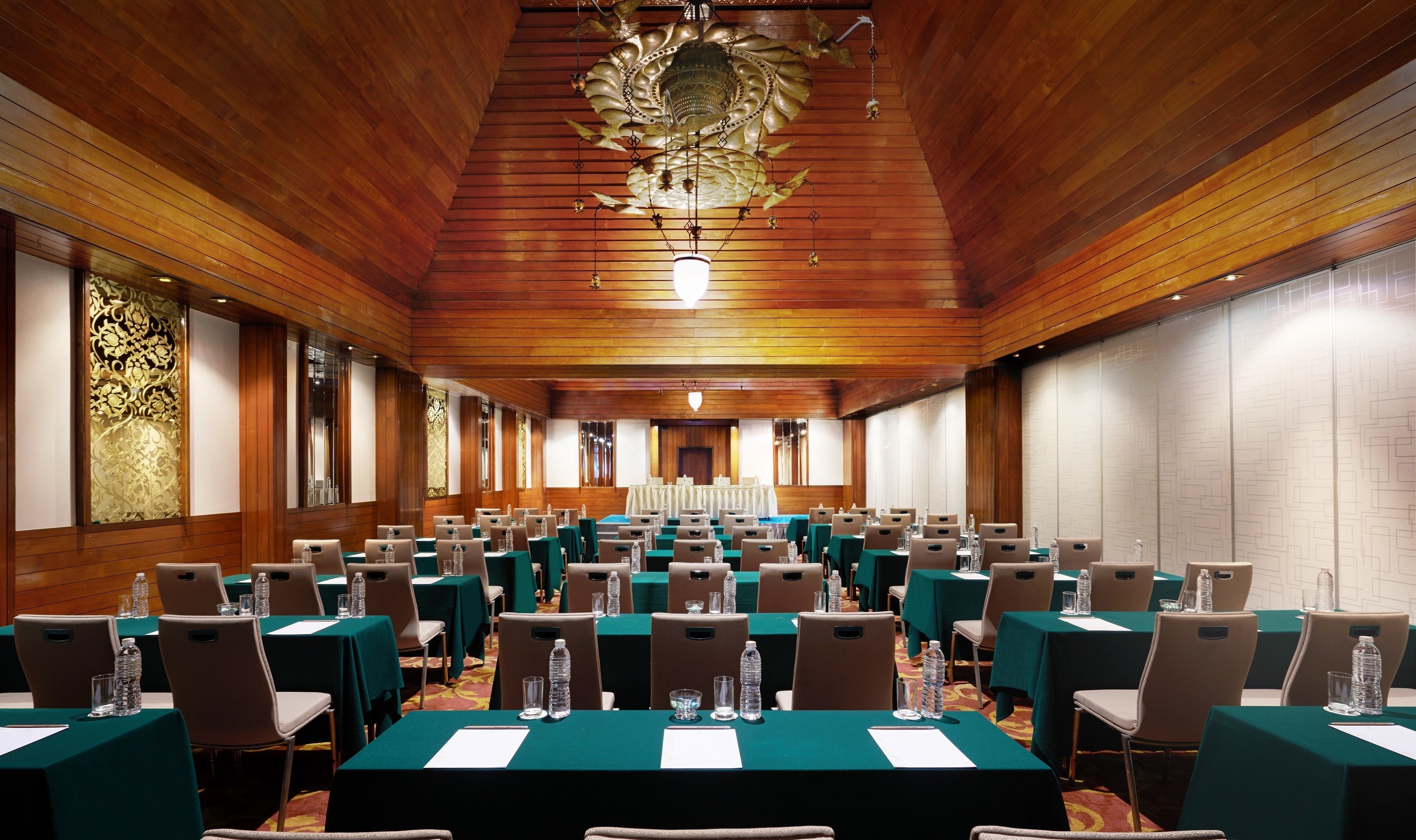 Uma sala de conferências com mesas e cadeiras dispostas em fileiras, painéis de madeira nas paredes e um lustre decorativo no teto.