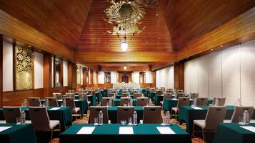 Uma sala de conferências com mesas e cadeiras dispostas em fileiras, painéis de madeira nas paredes e um lustre decorativo no teto.