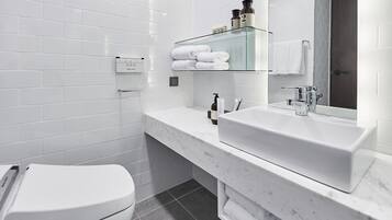 Uma casa de banho moderna com uma sanita branca, uma prateleira de vidro com artigos de higiene e uma grande pia com espelho acima.