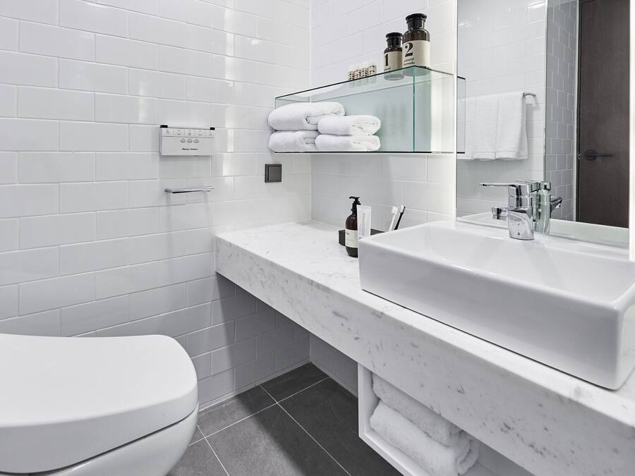 Uma casa de banho moderna com uma sanita branca, uma prateleira de vidro com artigos de higiene e uma grande pia com espelho acima.
