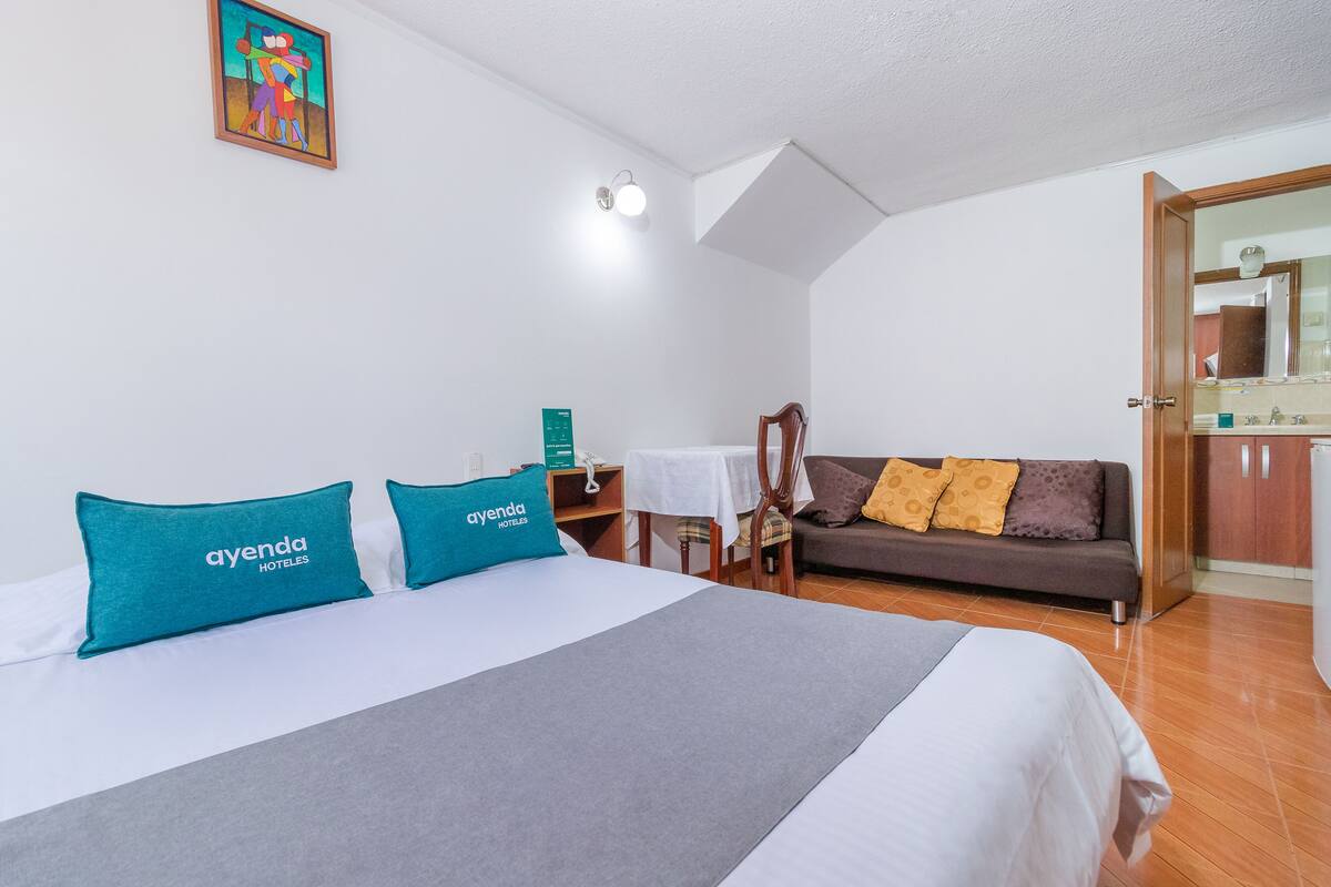 Quarto de hotel com cama, sofá, mesa com cadeira e pequeno armário.