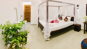 Quarto com cama de dossel, mesa de cabeceira com abajur, cadeira, pequena mesa com um vaso de flores e uma grande planta em vaso.
