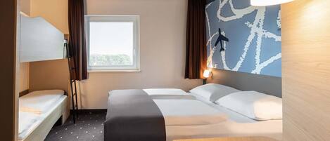 Quarto de hotel com uma cama beliche, uma cama de solteiro, uma escrivaninha e uma janela grande.