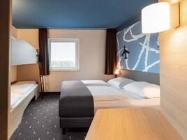 Quarto de hotel com uma cama beliche, uma cama de solteiro, uma escrivaninha e uma janela grande.
