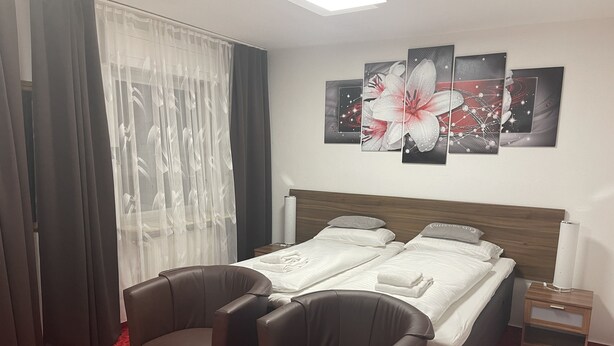 Quarto de hotel com duas camas, uma janela grande com cortinas leves, um tapete vermelho e uma parede com três obras de arte emolduradas.