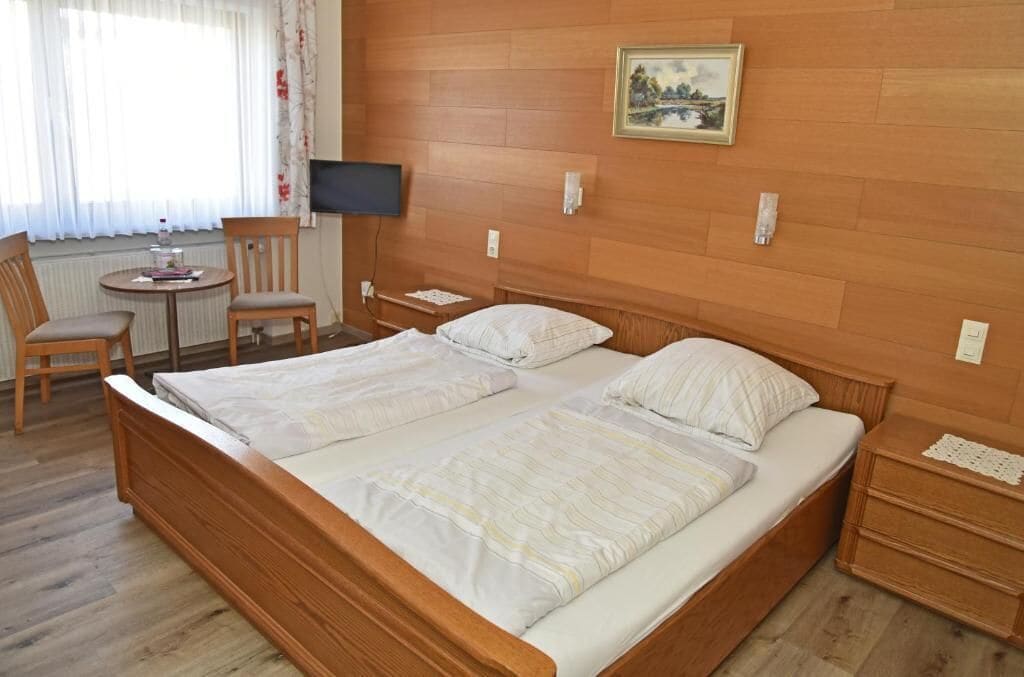 Uma cama de casal com cabeceira de madeira, uma mesa de cabeceira, uma cadeira, uma mesinha com um livro e uma garrafa, e uma TV fixada na parede.