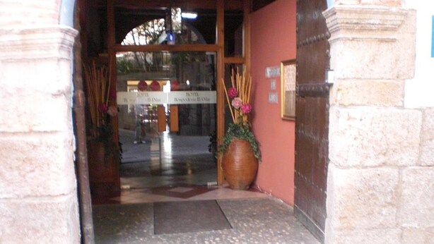 Entrada de hotel com porta de vidro, um vaso grande com flores e uma placa indicando o nome do hotel.
