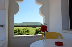 Mesa com um vaso de flores vermelhas e cinzeiro vermelho, uma cadeira amarela, e janela com vista para campos verdes e colinas.