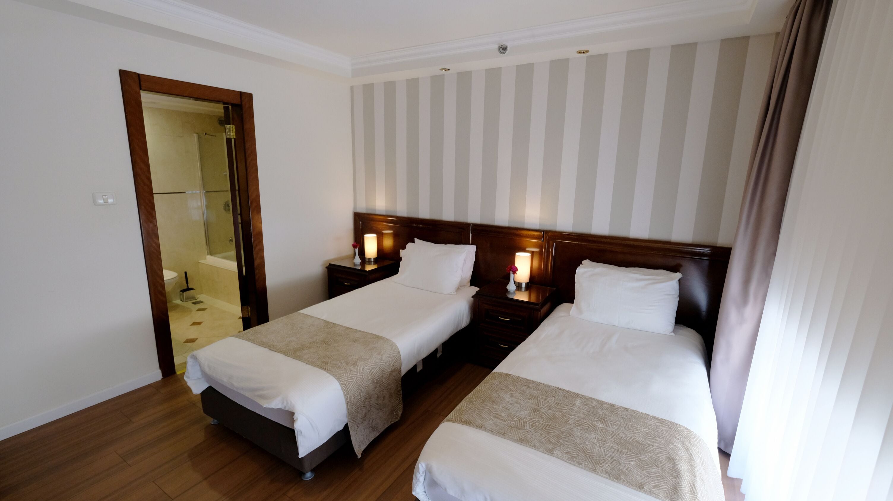 Quarto de hotel com duas camas, piso de madeira, área de chuveiro e papel de parede listrado.