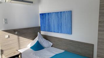 Quarto de hotel moderno com cabeceira de madeira, cama com roupa de cama branca e azul, um quadro abstrato azul na parede e uma pequena mesa de cabeceira com um abajur.