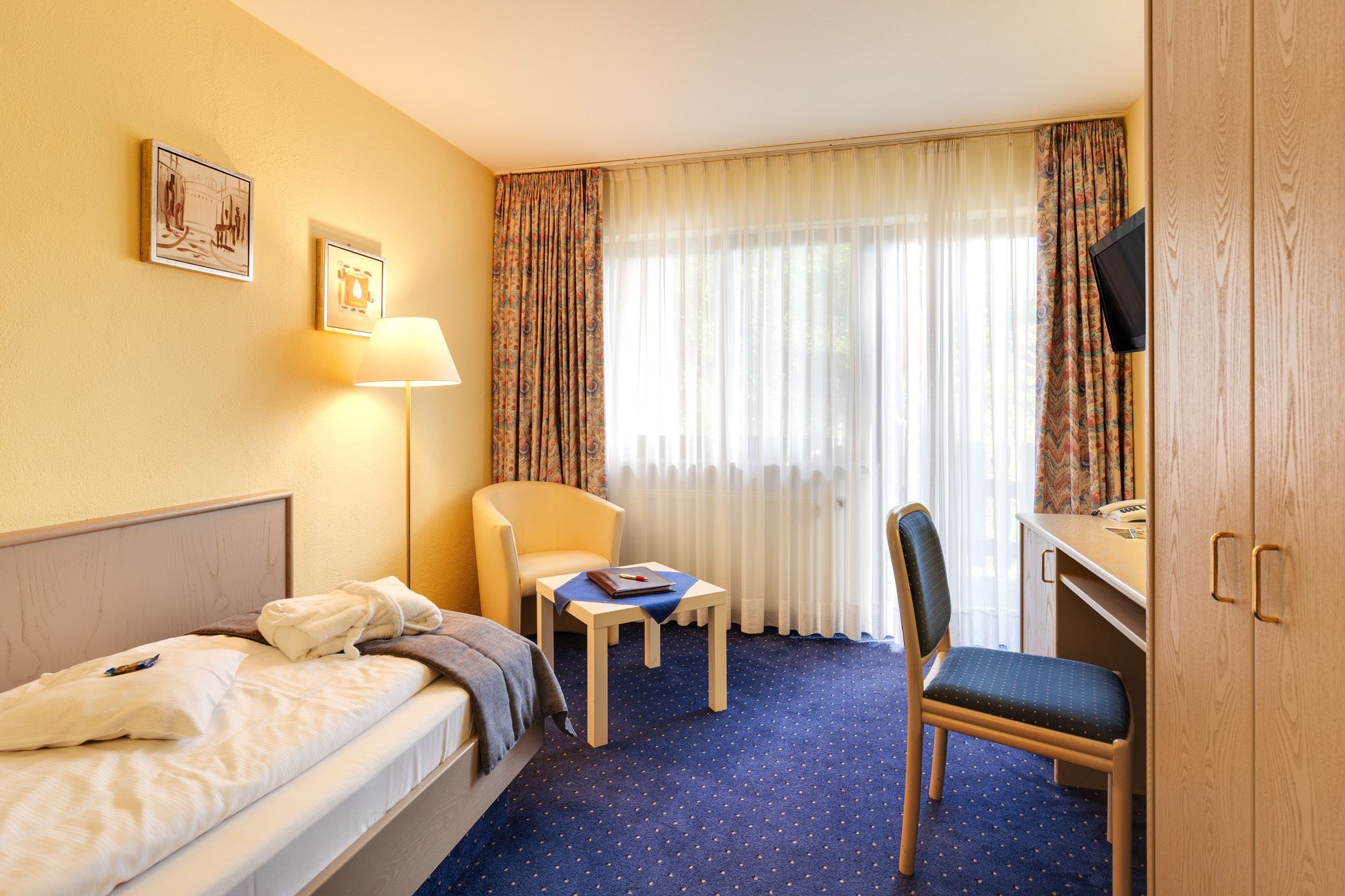 Quarto de hotel com cama, escrivaninha, cadeira, abajur, televisão e cortinas.