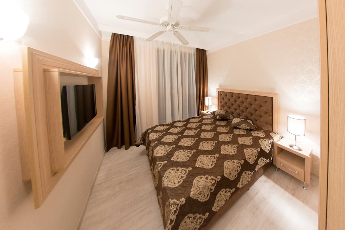 Quarto de hotel com cama, televisão fixada na parede, mesa de cabeceira com abajur e ventilador de teto.