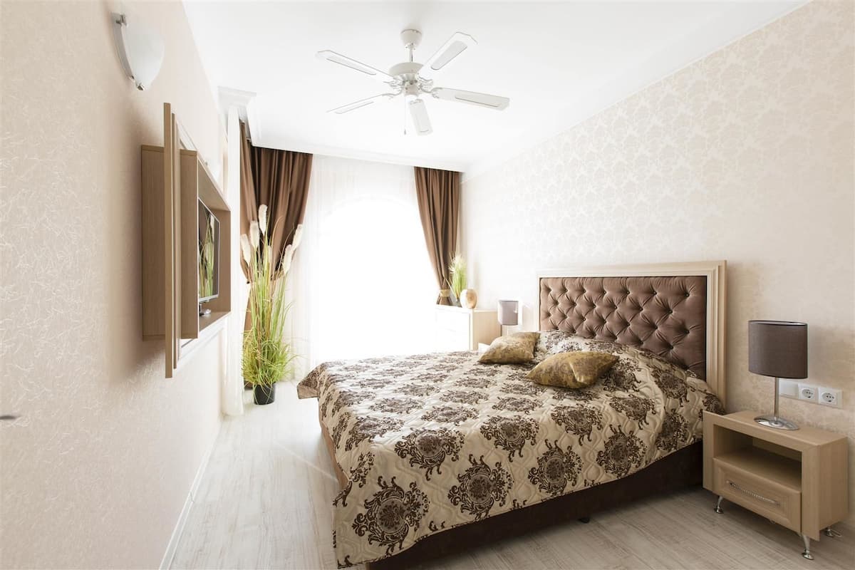 Quarto com cabeceira estofada, mesa de cabeceira com abajur, mesa de apoio e planta em vaso.