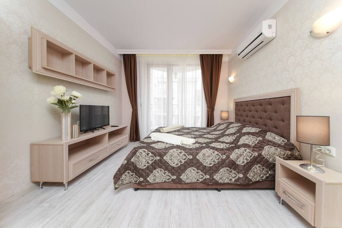 Quarto com uma cama grande, uma TV sobre um suporte de madeira, uma mesa de cabeceira com abajur e um aparelho de ar condicionado na parede.