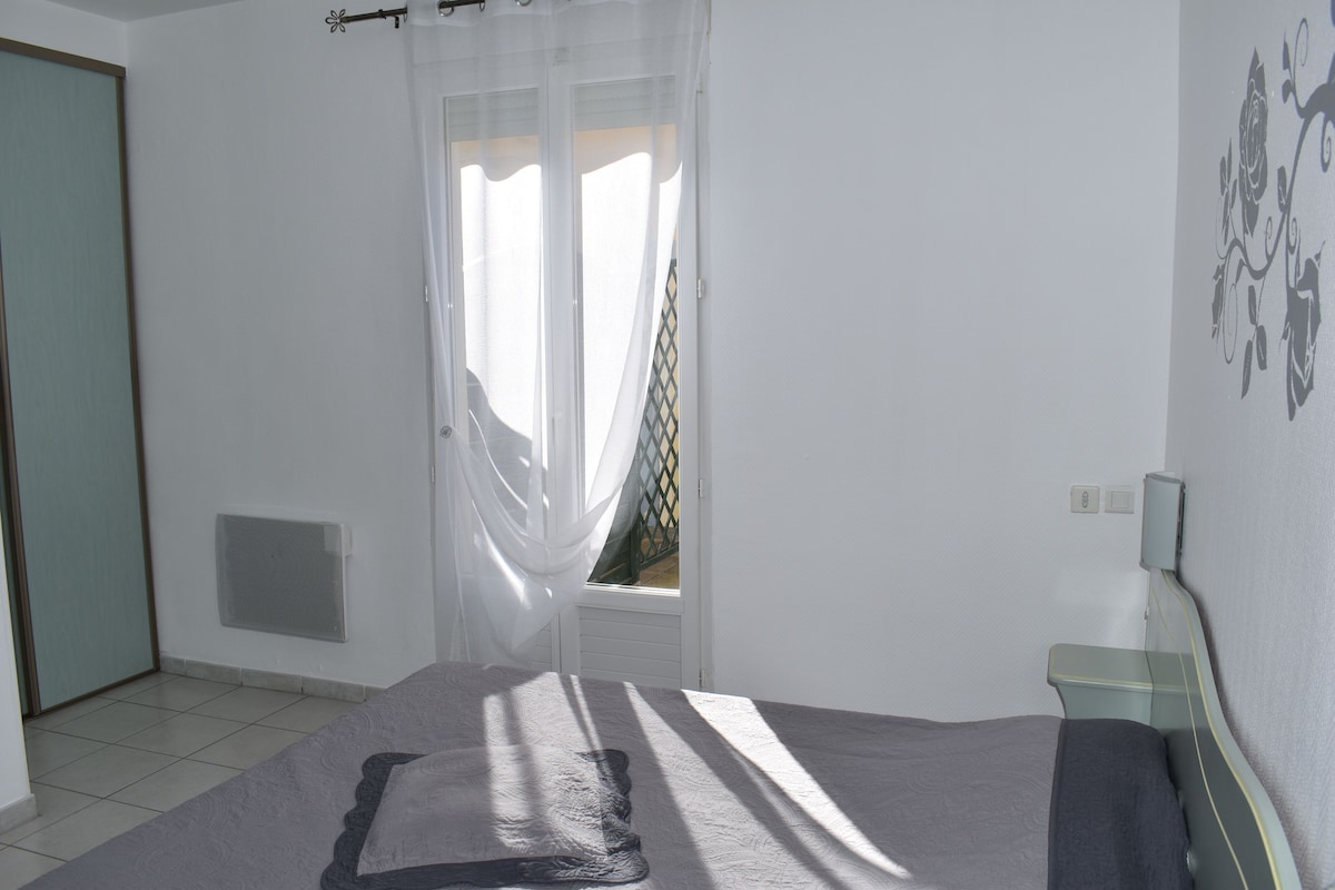 Quarto com cama, janela com cortinas leves, luminária na parede e uma pequena mesa de cabeceira.
