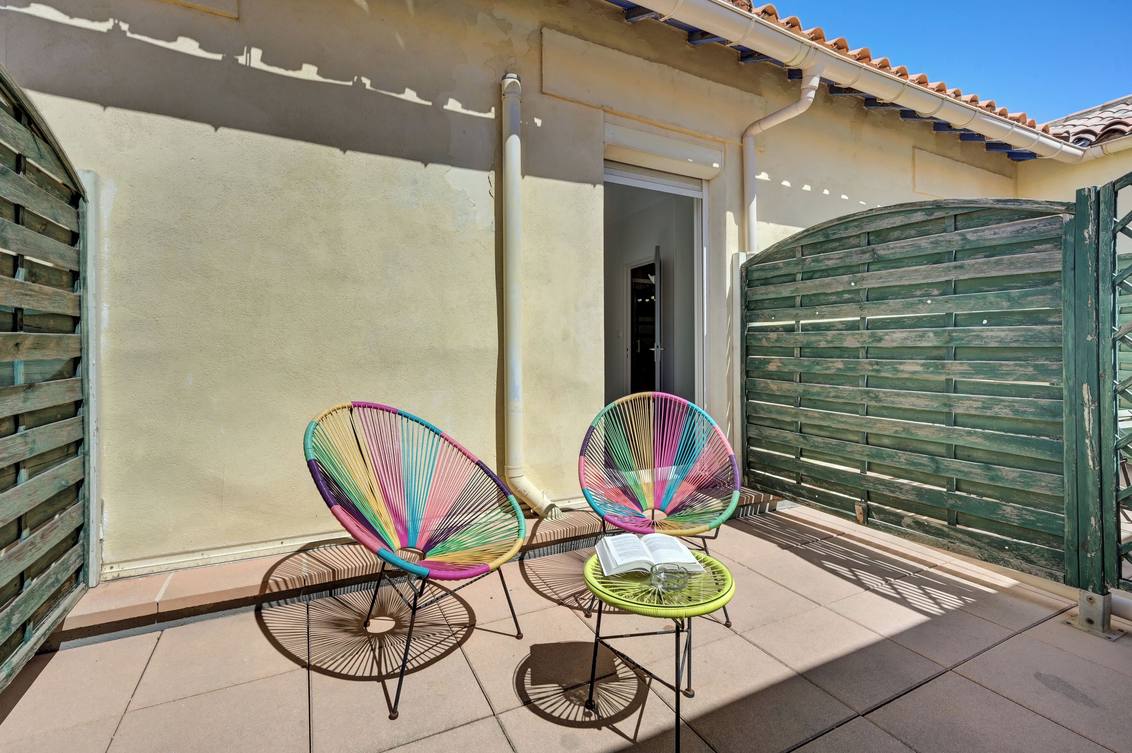 Um pátio com duas cadeiras coloridas e uma mesa pequena, um portão verde e uma parede bege.