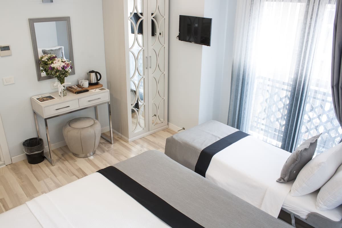 Quarto de hotel com duas camas, uma escrivaninha com cadeira, um banquinho pequeno, um cesto de lixo, um espelho, uma TV e uma janela com cortinas.
