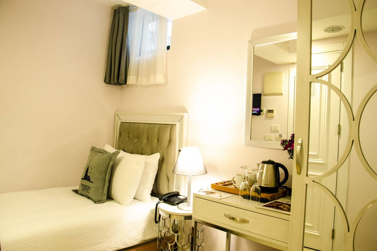 Quarto de hotel com cama, mesa de cabeceira com abajur, telefone e bandeja com cafeteira e xícaras.