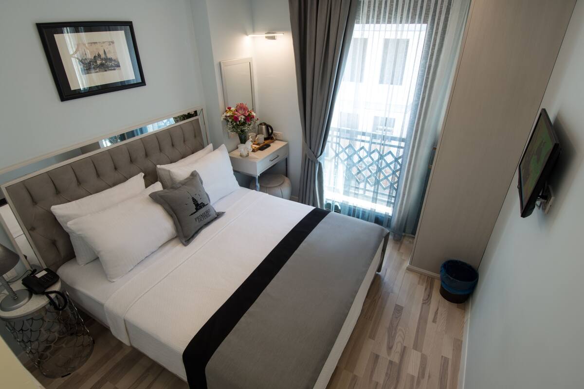 Quarto de hotel com uma cama grande, uma mesa de cabeceira com abajur, um espelho e uma janela com cortinas.