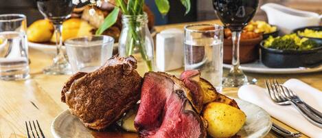 Um prato de carne com acompanhamento de batatas e um copo de vinho tinto sobre uma mesa de madeira.