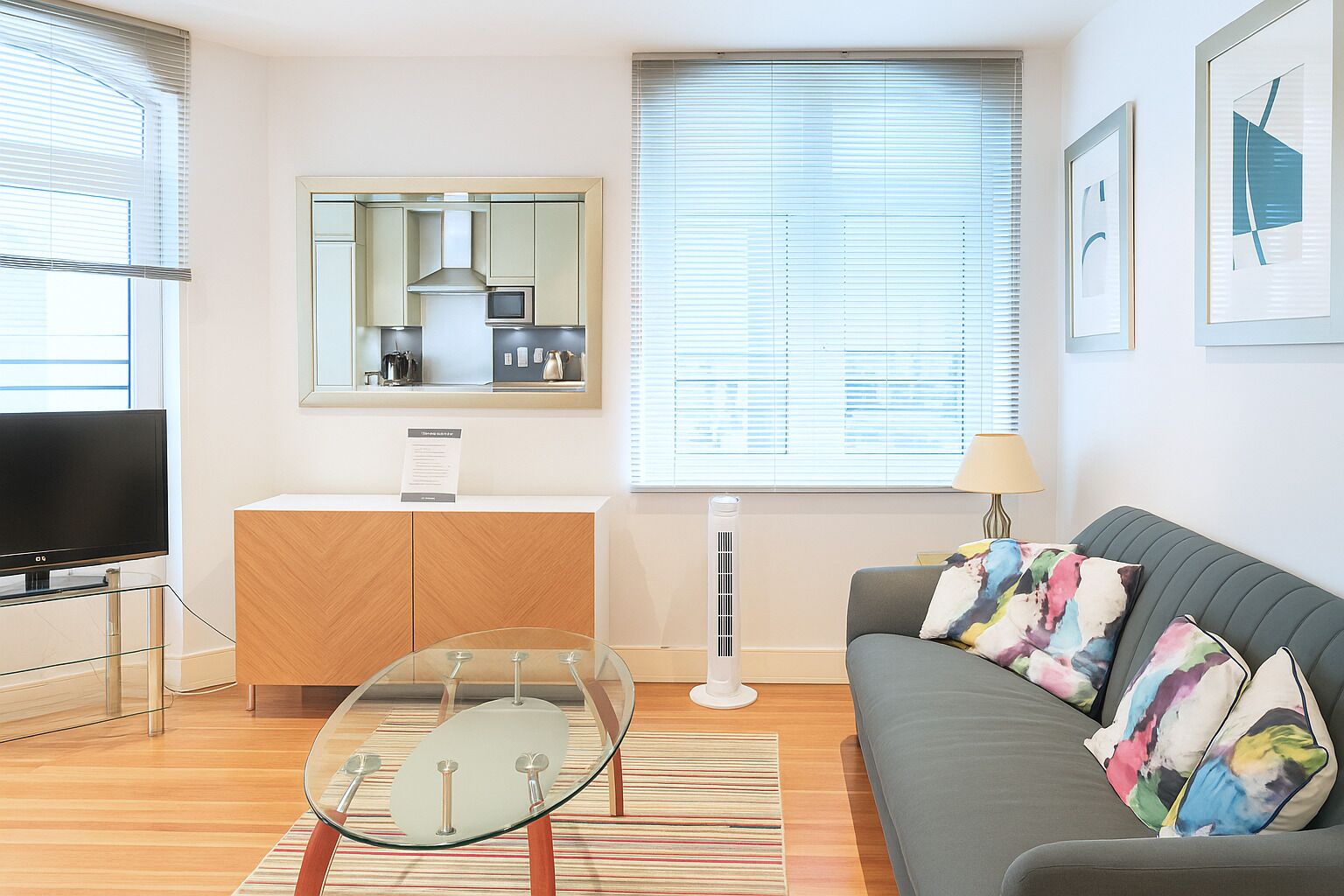 Uma sala de estar moderna com uma mesa de centro de vidro, um sofá cinzento com almofadas coloridas, uma televisão, uma área de cozinha com micro-ondas e uma grande janela com persianas.