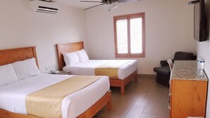 Quarto de hotel com duas camas de madeira, um ventilador de teto, uma janela com persianas, uma pequena escrivaninha com cadeira e uma TV fixada na parede.