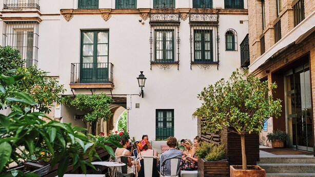 Um café ao ar livre com mesas e cadeiras, rodeado por edifícios com portões verdes e fachada branca.