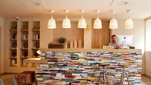 Uma mesa de recepção com uma pilha de livros atrás, prateleiras de madeira à esquerda e um homem trabalhando no balcão.