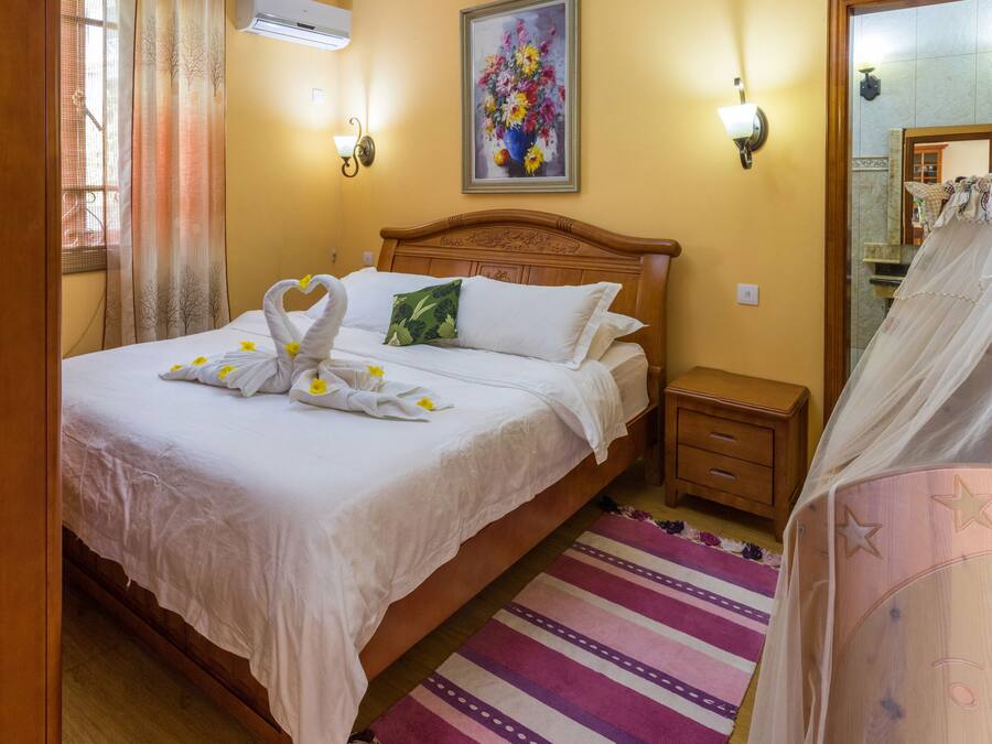 Cama bem arrumada com lençóis brancos e toalhas decorativas em formato de cisnes, cabeceira de madeira, mesa de cabeceira pequena, luminária fixada na parede e um quadro com flores.