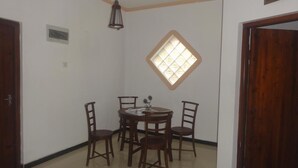 Uma pequena área de refeições com uma mesa redonda e quatro cadeiras, uma janela com padrão de losangos e um lustre.