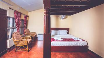 Quarto de hotel com cama de madeira, duas cadeiras de vime, uma mesinha e uma janela com cortinas.