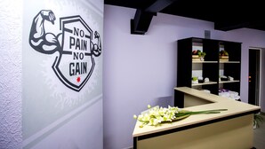 Uma área de recepção com um letreiro que diz 'NO PAIN NO GAIN', um balcão de recepção e uma prateleira com toalhas e produtos de higiene pessoal.