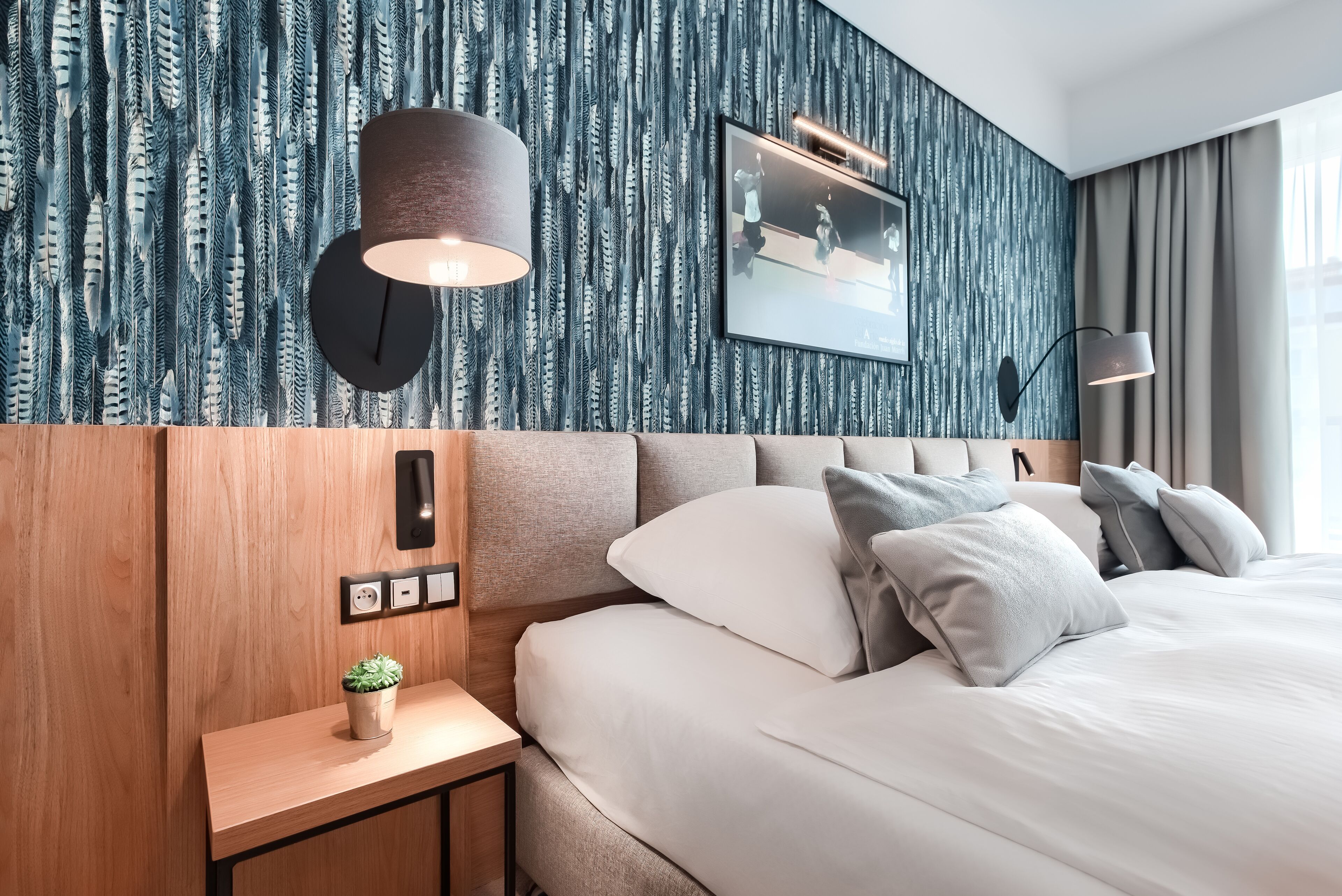 Quarto de hotel moderno com cabeceira de madeira, cama grande com várias almofadas, mesa de cabeceira pequena com uma planta e televisão na parede.