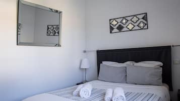 Quarto de hotel com cama, mesa de cabeceira, luminária, espelho e um objeto decorativo na parede.
