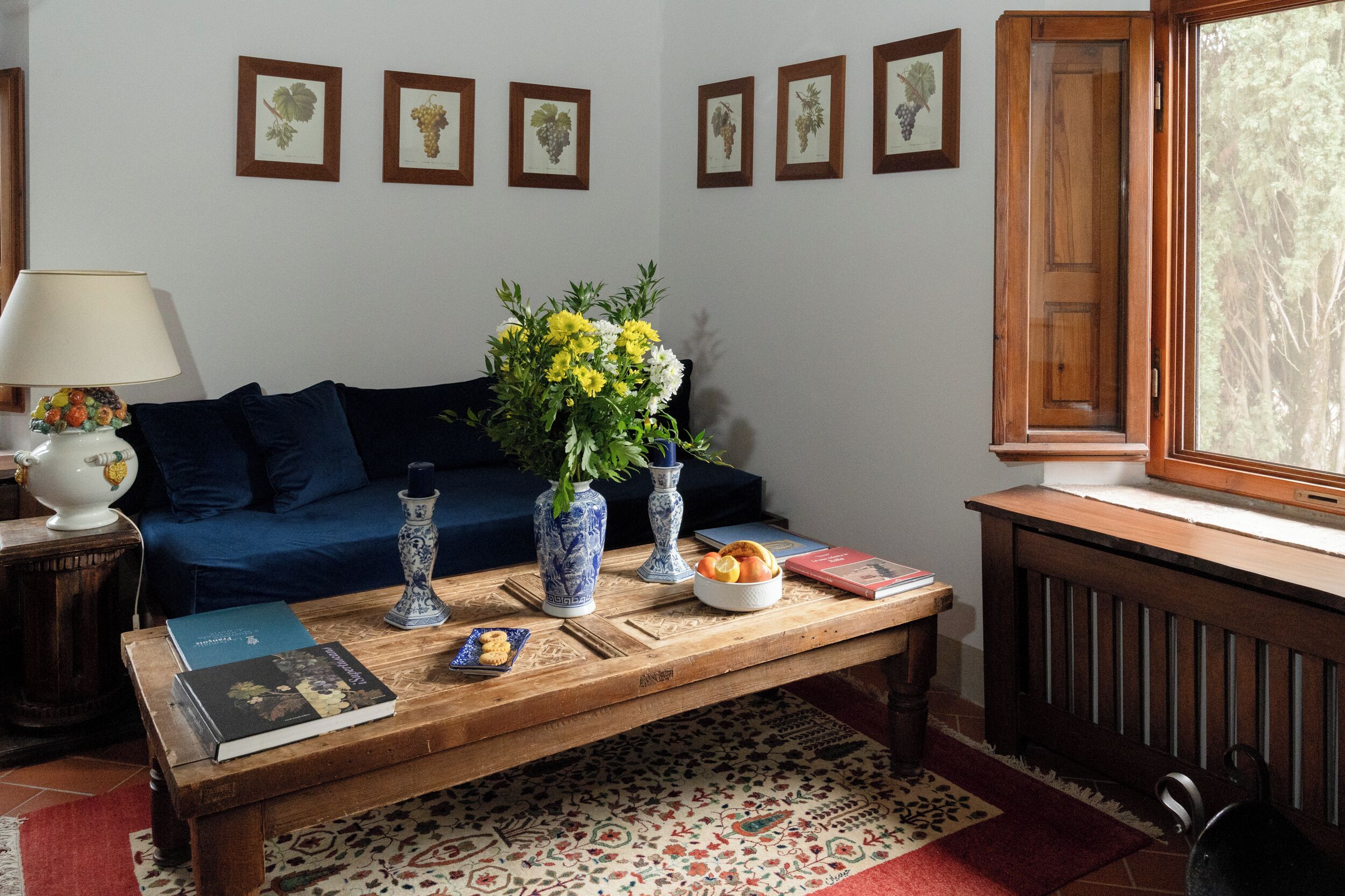 Uma sala de estar com um sofá azul, uma mesa de centro de madeira com livros e objetos decorativos, um vaso com flores e quadros nas paredes.