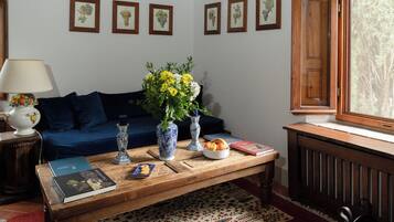 Uma sala de estar com um sofá azul, uma mesa de centro de madeira com livros e objetos decorativos, um vaso com flores e quadros nas paredes.