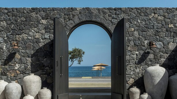 Abre-se uma porta para uma praia, com paredes de pedra e grandes vasos.